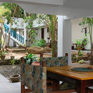 Uma mesa de madeira com cadeiras estampadas, rodeada por plantas em vasos e uma escada com grades azuis que leva a um edifício branco.