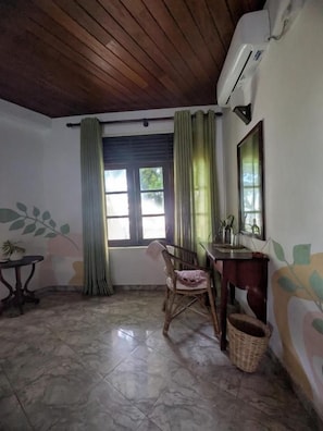 Quarto com teto de madeira, janela com cortinas, uma cadeira, uma mesa e espelho fixado na parede.