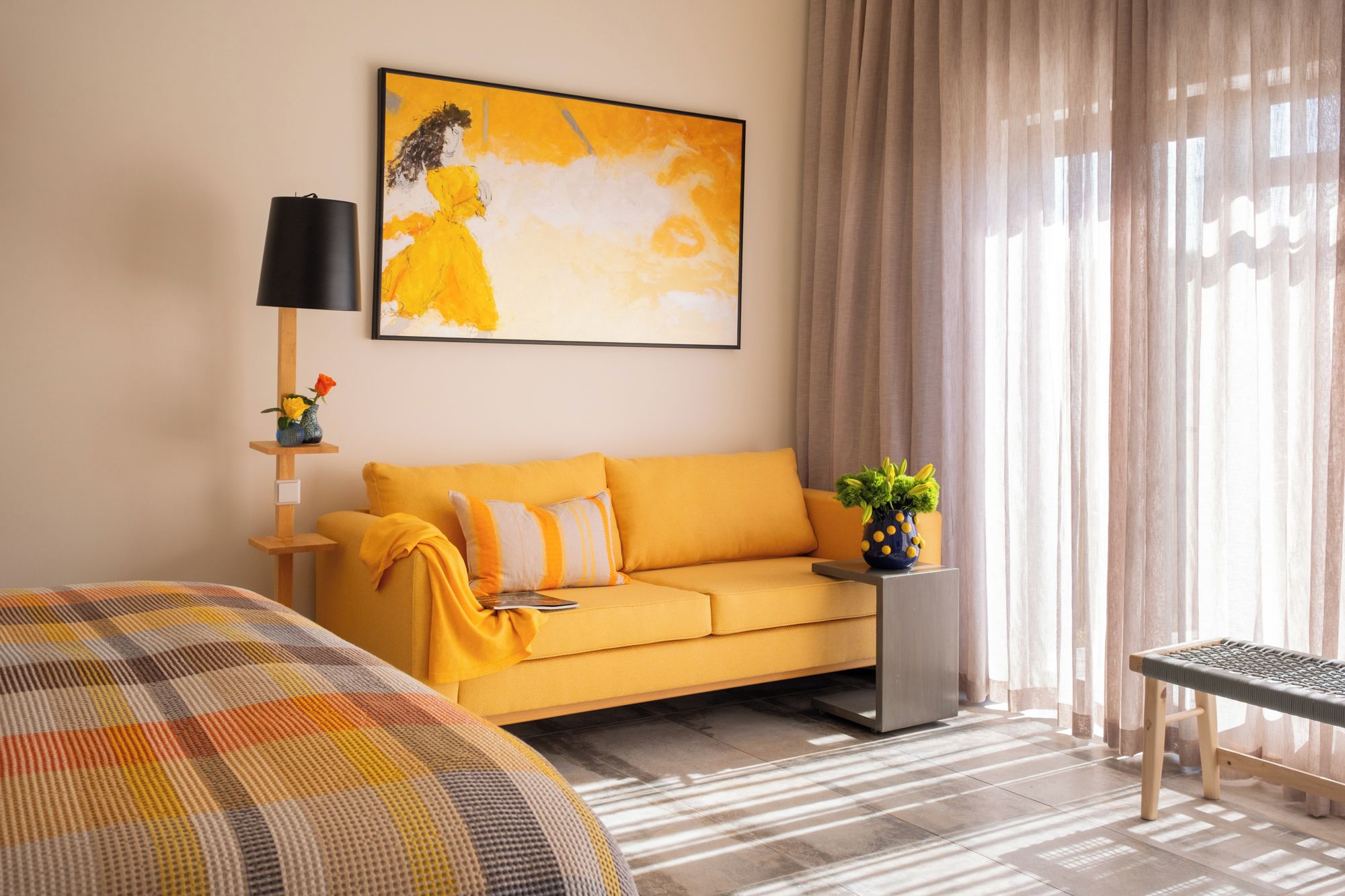 Quarto de hotel com sofá, mesa de cabeceira com luminária, pintura na parede e janela com cortinas.