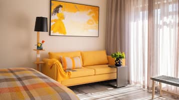 Quarto de hotel com sofá, mesa de cabeceira com luminária, pintura na parede e janela com cortinas.