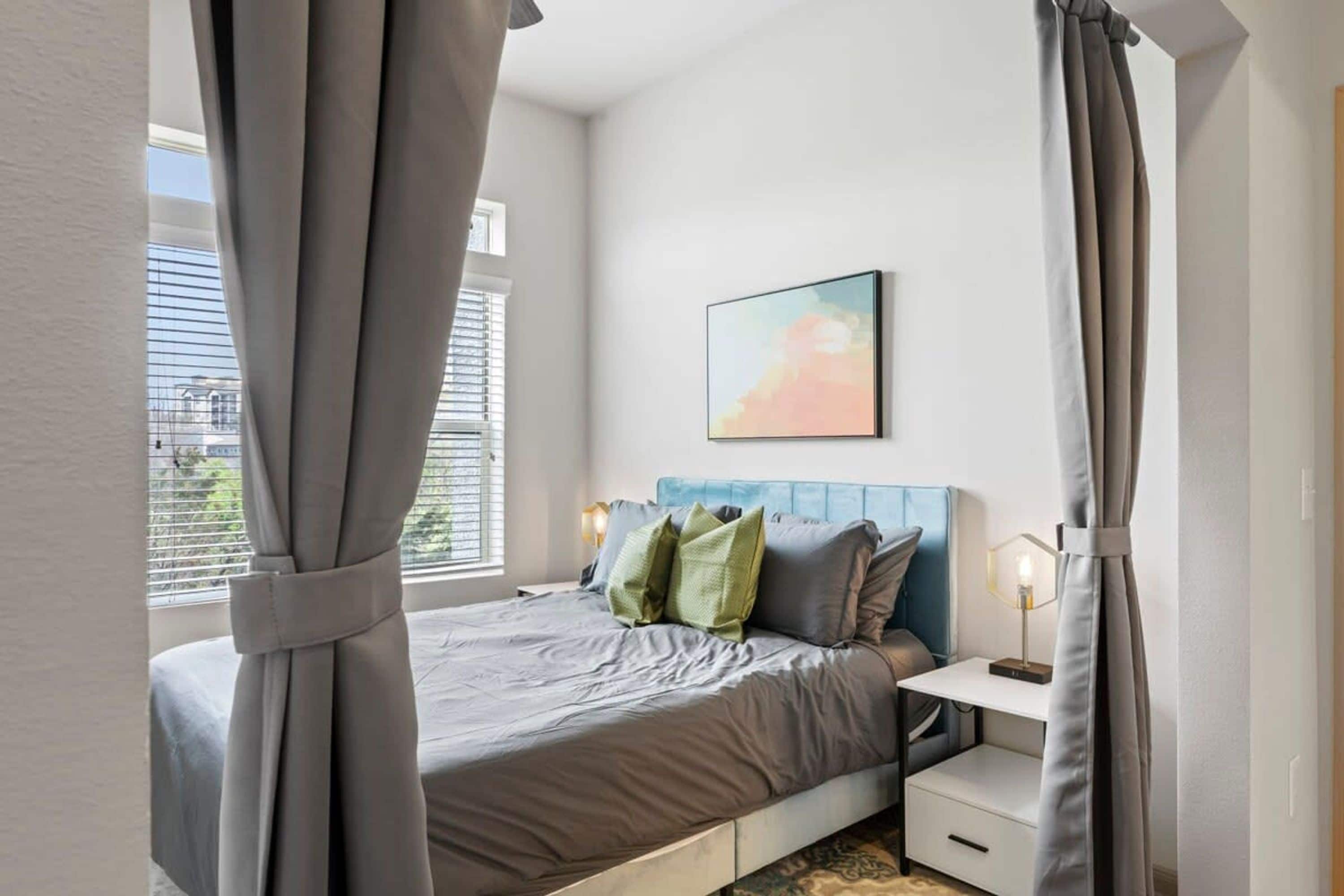 Quarto com cama, almofadas, mesa de cabeceira, abajur e um quadro na parede.