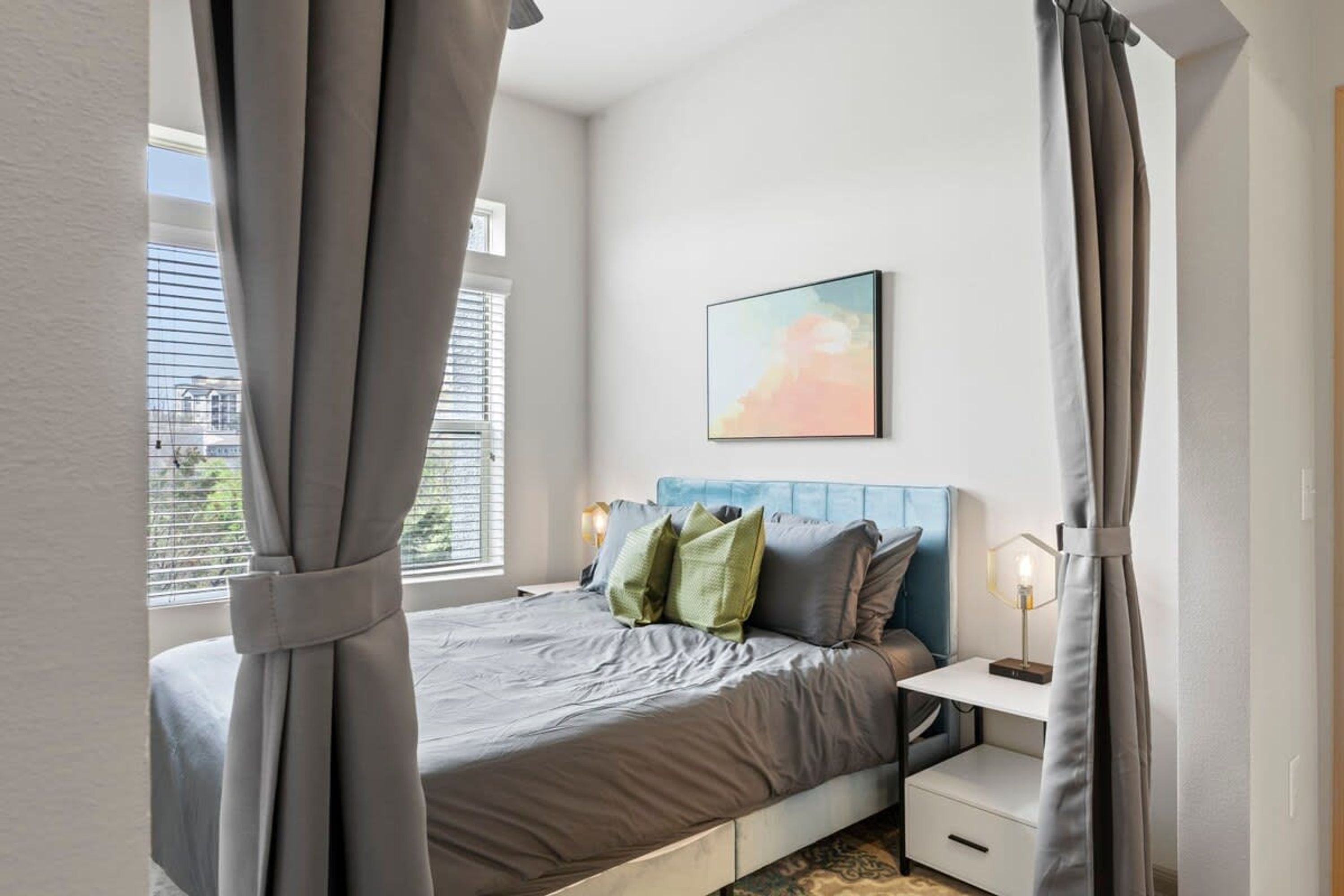 Quarto com cama, almofadas, mesa de cabeceira, abajur e um quadro na parede.