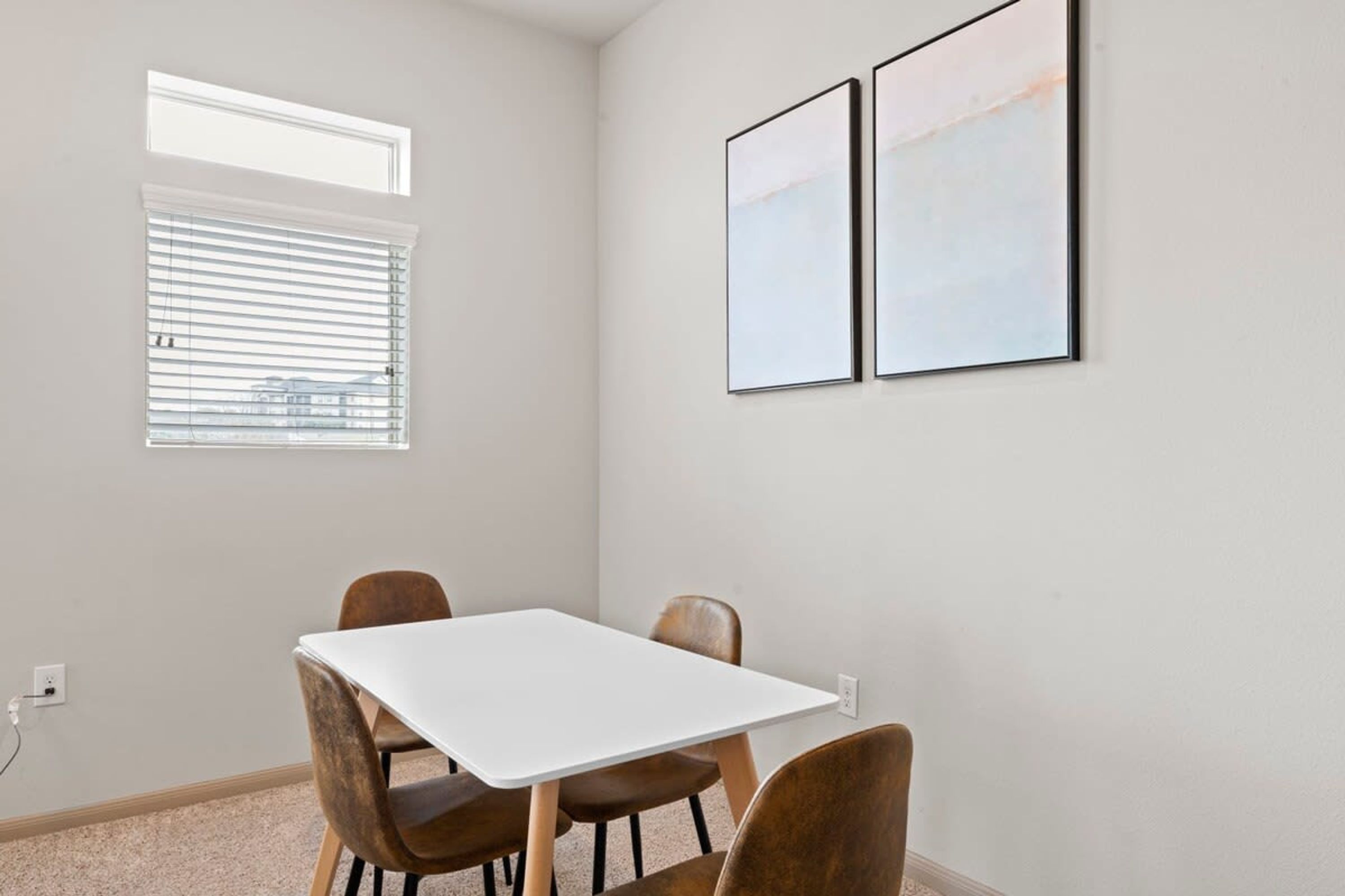 Quarto com uma mesa branca e quatro cadeiras, uma janela com persianas e duas imagens emolduradas na parede.
