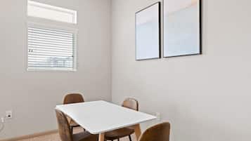 Quarto com uma mesa branca e quatro cadeiras, uma janela com persianas e duas imagens emolduradas na parede.