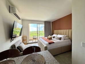 Quarto de hotel com cama, televisão, varanda com cadeira e vista para o verde.