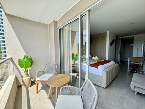 Varanda com mesa e cadeiras, um vaso de planta e porta de vidro deslizante que dá para um quarto com cama e cômoda.