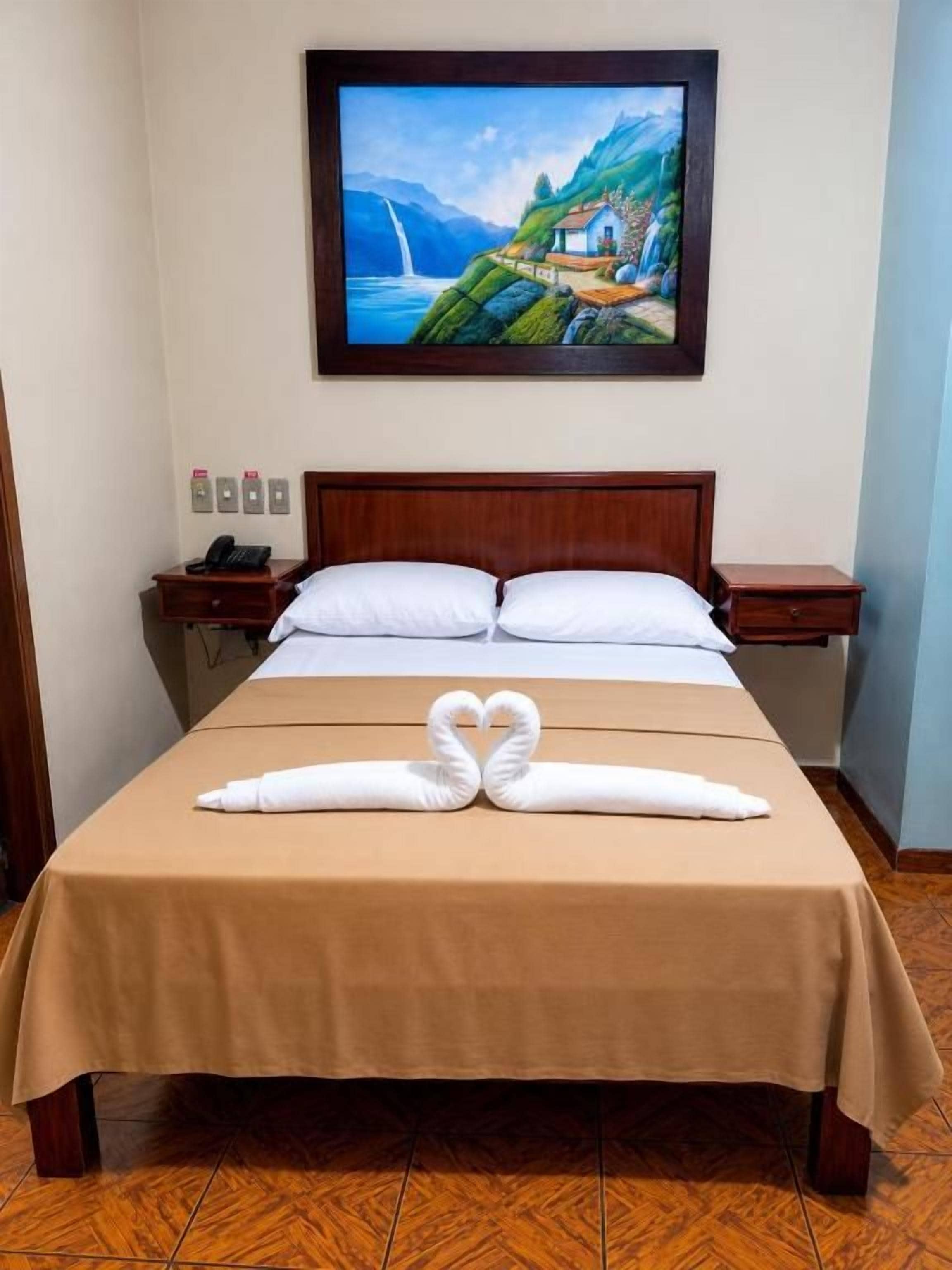 Quarto de hotel com uma cama, um quadro de uma paisagem e uma toalha dobrada em formato de coração sobre a cama.