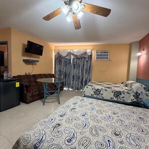 Quarto de hotel com cama, sofá, mesinha, ventilador de teto, televisão e aparelho de ar condicionado.