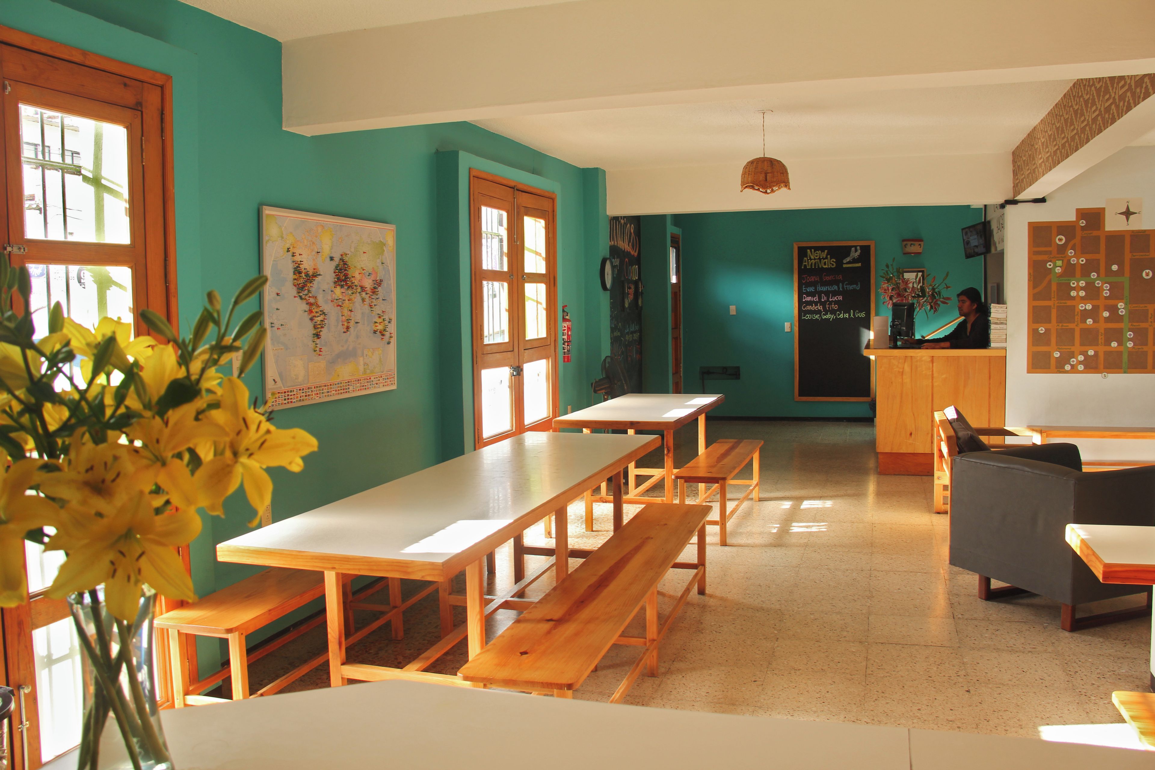 Um quarto com mesas e bancos de madeira, um quadro-negro com um mapa e um mural com uma lista de nomes.