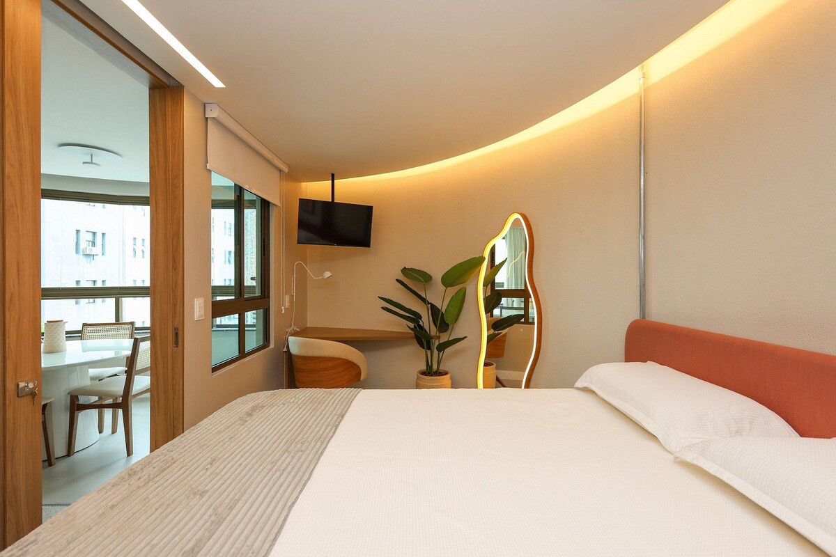 Quarto de hotel com cama, escrivaninha, cadeira, planta em vaso, espelho e televisão.