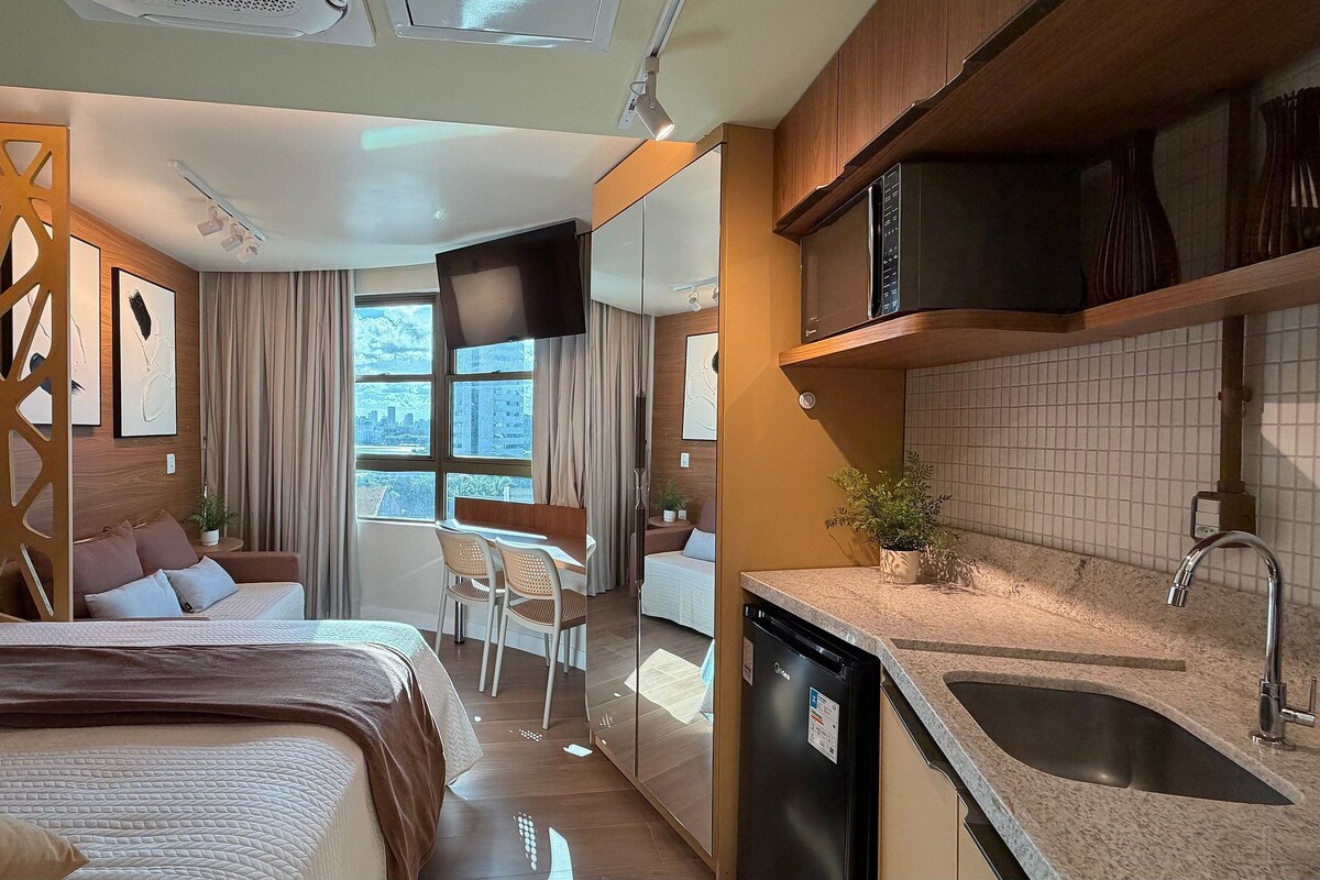 Quarto de hotel compacto com cama, escrivaninha, cadeira, pequeno espaço de cozinha e televisão.