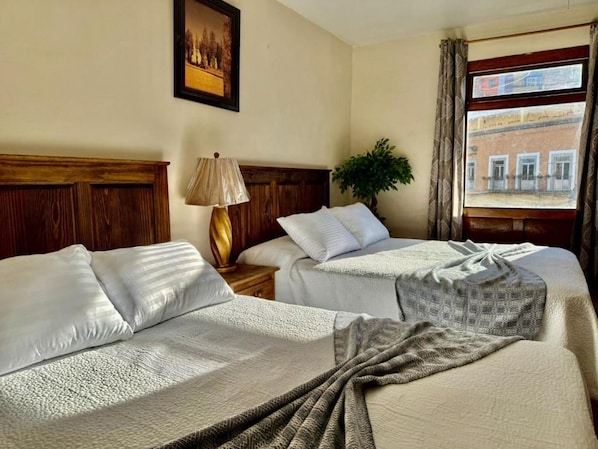 Quarto de hotel com duas camas, uma mesa de cabeceira, abajur, planta e janela com vista para prédios.