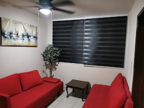 Uma sala de estar com dois sofás vermelhos, uma mesa pequena, um ventilador de teto, um quadro e uma janela com persianas.