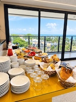 Um buffet de café da manhã com diversos alimentos, utensílios, uma garrafa de ketchup e vista para o oceano através de grandes janelas.