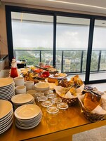 Uma mesa de buffet com diversos alimentos e pratos empilhados, com o cenário de uma varanda e vista para edifícios e vegetação.