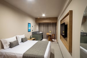 Quarto de hotel com cama, escrivaninha, cadeira, televisão e um espelho grande.