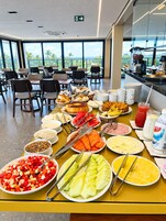 Buffet montado com diversos alimentos e utensílios, área de refeições com cadeiras e mesas, e vista para o exterior através de amplas janelas.