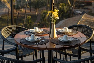 Mesa de madeira redonda posta para quatro, com jogos americanos, xícaras brancas e um vaso com flores amarelas.