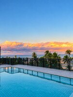 Piscina com vista para o oceano e palmeiras ao pôr do sol.