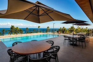 Área da piscina com vista para o oceano, mobiliário de exterior e guarda-sóis.
