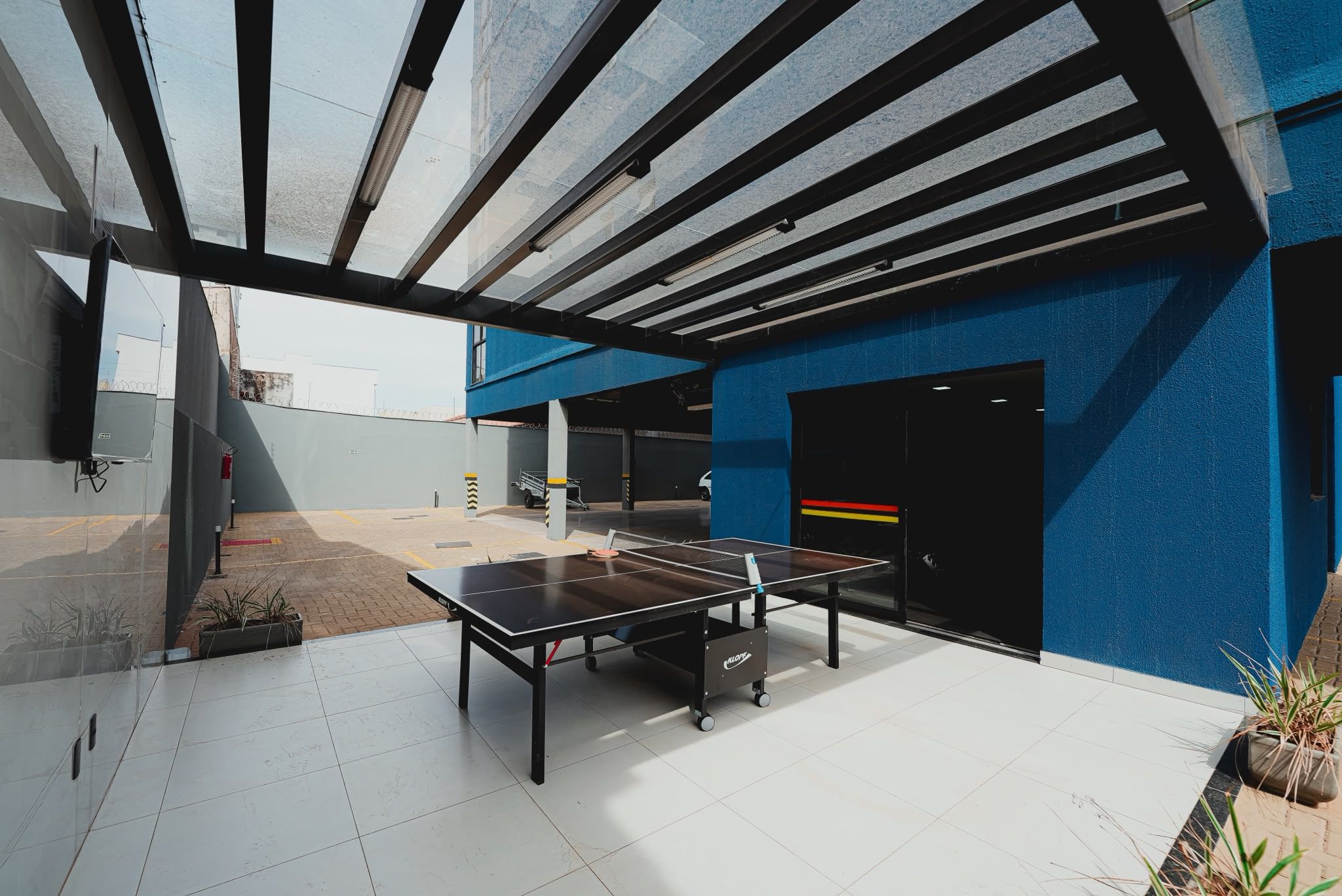 Mesa de pingue-pongue ao ar livre instalada em área coberta, com parede azul e entrada de garagem.