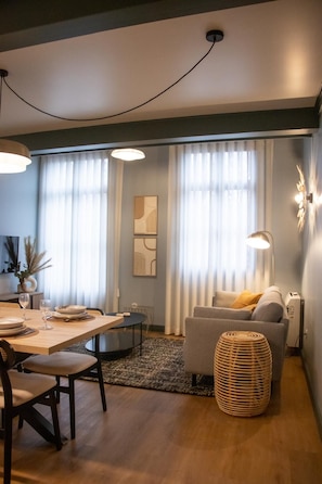 Uma sala de estar moderna com área de jantar, apresentando um sofá, uma mesa redonda e uma janela grande com cortinas.