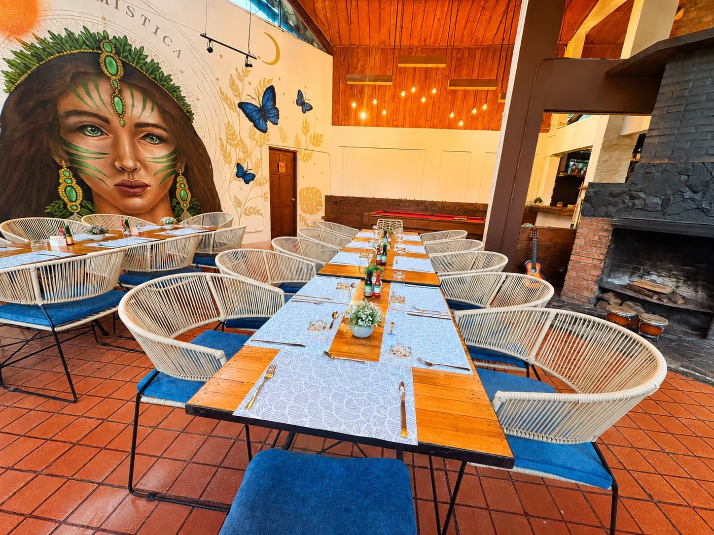 Um restaurante com mesas grandes e compartilhadas, cadeiras de vime e um mural na parede.