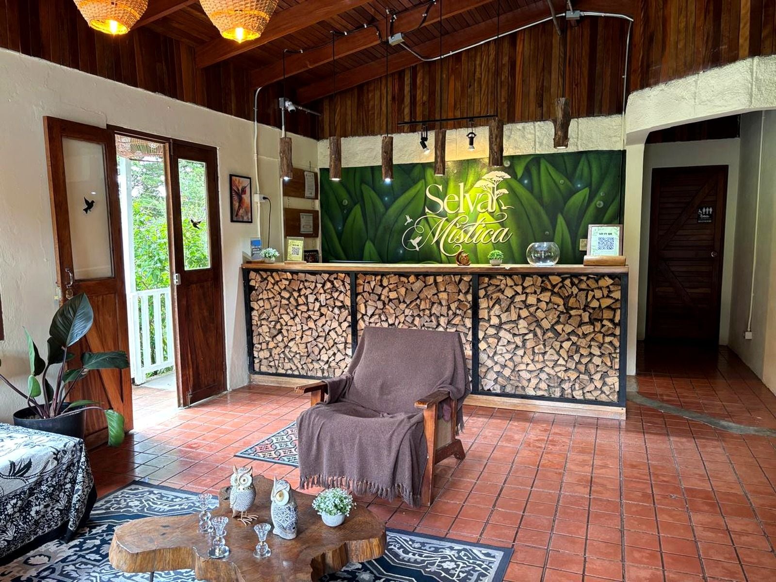 Área de recepção com teto de madeira, balcão de atendimento com fundo verde e área de estar com uma cadeira e uma mesa.