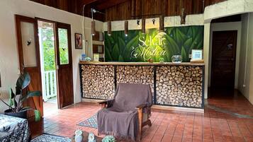 Área de recepção com teto de madeira, balcão de atendimento com fundo verde e área de estar com uma cadeira e uma mesa.