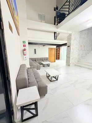 Um hall amplo com um sofá modular cinza, uma pequena mesa branca, uma escadaria e uma parede de mármore.