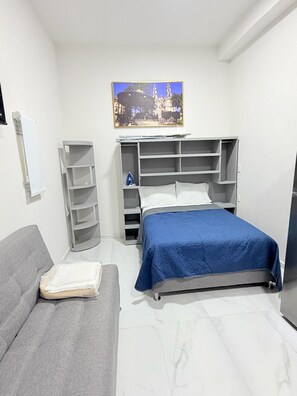 Quarto com cama, sofá cinzento, televisão na parede e estante de livros.