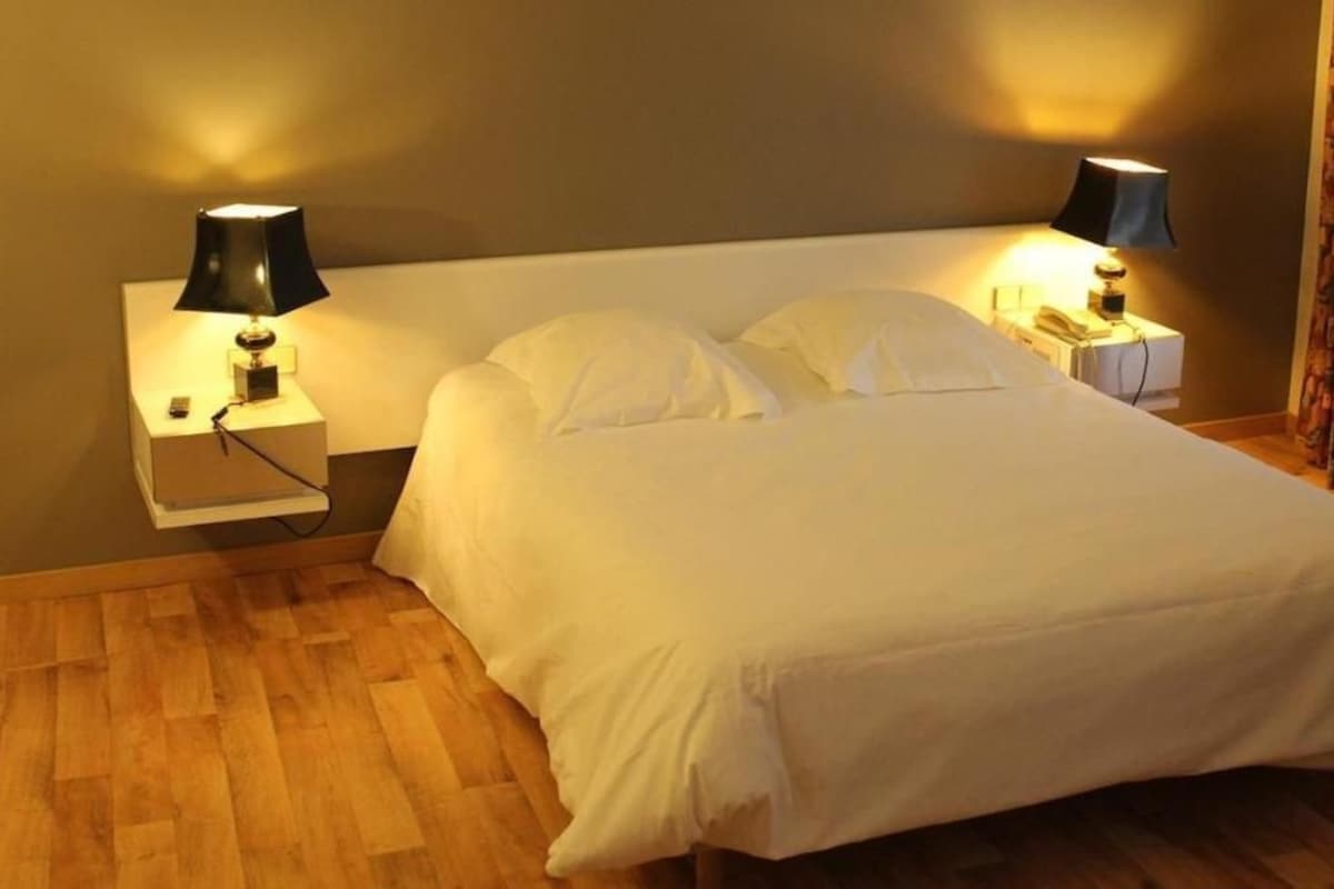 Cama bem arrumada com roupa de cama branca, duas luminárias de cabeceira e piso de madeira.