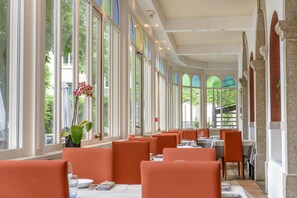 Um restaurante com janelas altas, assentos laranja e vista para o verde.