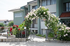 Um edifício com uma placa indicando 'GARDENIA' e um arco decorativo coberto de rosas trepadeiras.