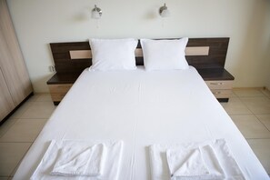 Cama de casal com lençóis brancos, duas mesas de cabeceira e luminárias fixadas na parede.