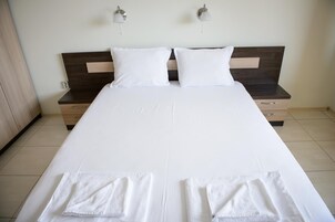 Cama de casal com lençóis brancos, duas mesas de cabeceira e luminárias fixadas na parede.