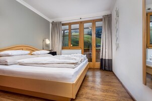 Quarto com cama de madeira, mesa de cabeceira, abajur e uma janela grande com cortinas.