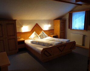 Quarto com cama de madeira, duas mesas de cabeceira, um criado-mudo, janela com cortinas e radiador.