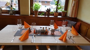 Mesa de jantar posta para seis pessoas, com taças de vinho, talheres e guardanapos laranja.