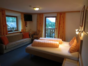 Quarto de hotel com sofá, cama com duas almofadas, mesa pequena, TV fixada na parede e varanda com cortinas.