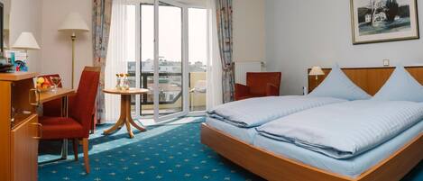 Quarto de hotel com cama, escrivaninha, cadeira, mesinha e vista da cidade pela janela.