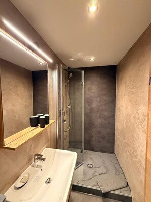 Uma casa de banho moderna com chuveiro em cabine de vidro, toucador com lavatório e prateleira com objetos cilíndricos pretos.