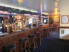Uma área de bar com banquetas, um balcão com diversos tipos de bebidas e uma parede com o menu.