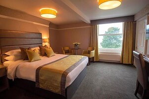 Quarto de hotel com uma cama grande, mesas de cabeceira, escrivaninha e janela com cortinas.