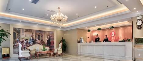 Um saguão de hotel com balcão de recepção, um candelabro, uma pintura grande e um padrão geométrico no chão.