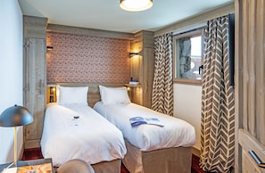 Quarto de hotel com duas camas de solteiro, cabeceira de madeira, parede estampada, janela com cortinas e mesa de cabeceira com abajur.