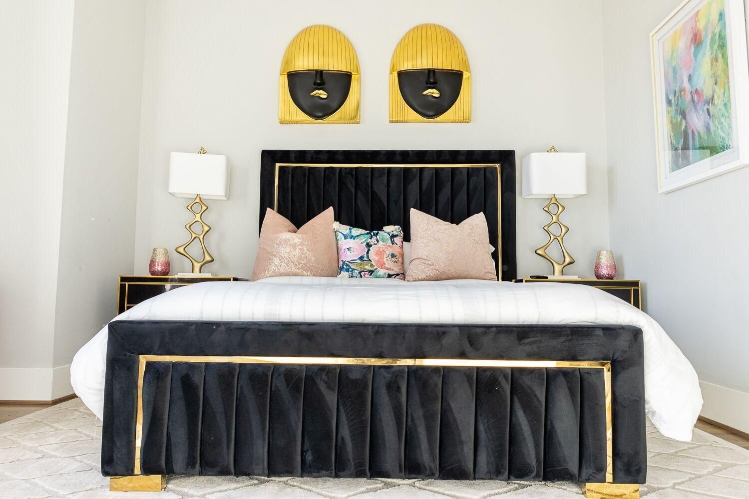 Uma cama com cabeceira preta, duas luminárias fixadas na parede e um quadro abstrato emoldurado.