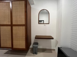 Quarto com guarda-roupa de madeira, prateleira pequena e lixeira, além de um espelho na parede.