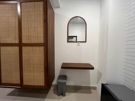 Quarto com guarda-roupa de madeira, prateleira pequena e lixeira, além de um espelho na parede.