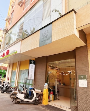 Um edifício com uma placa indicando 'MAXHILL INN' e uma farmácia Saúde. Motos estão estacionadas na frente, e uma placa mostra 'Quartos 3F Elevador e Ascensor'.